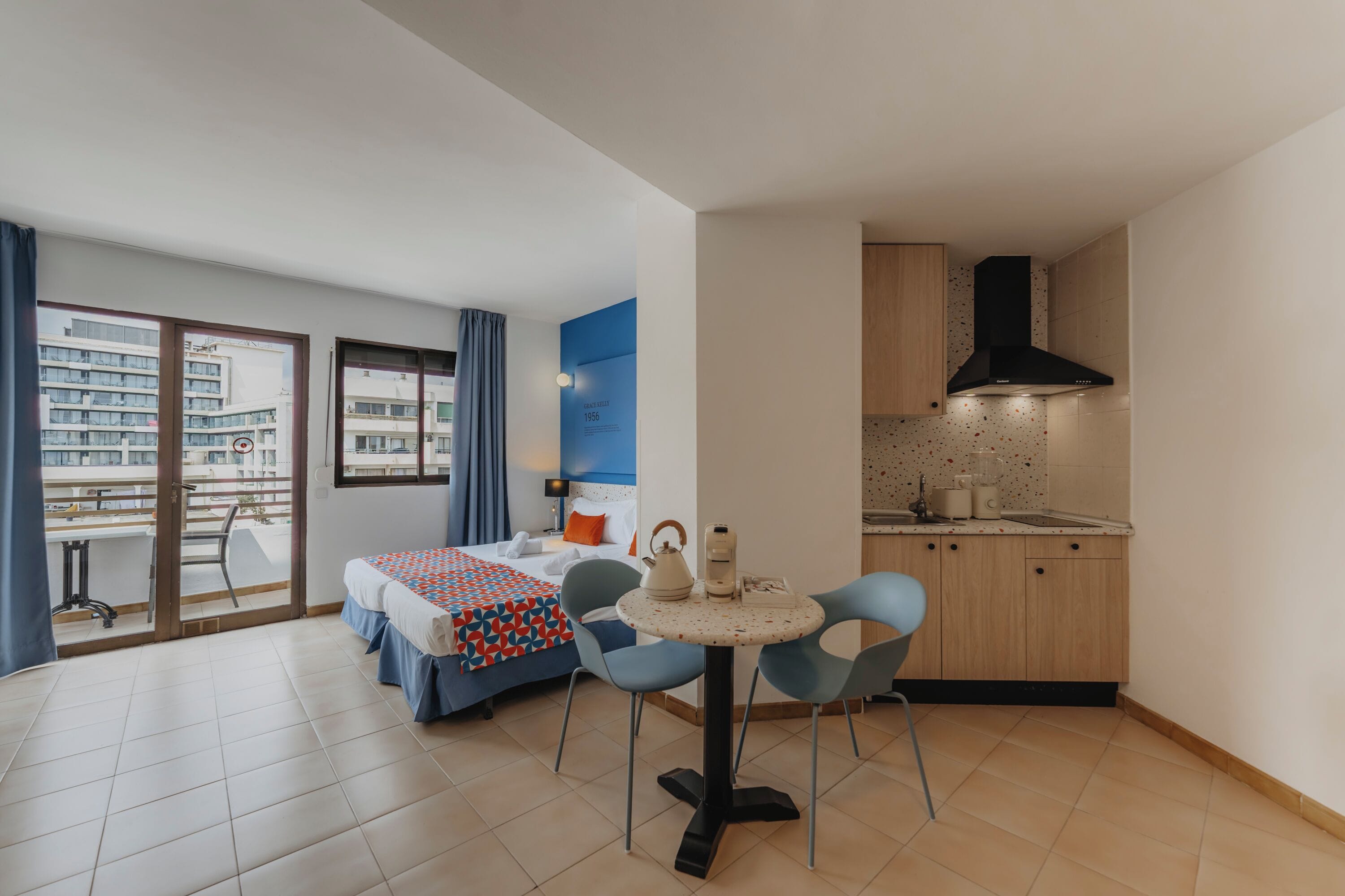Quarto de hotel moderno com cama, mesa de jantar com cadeiras, cozinha compacta e varanda com vista para edifícios.