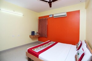 Quarto de hotel com cama, aparelho de ar condicionado, ventoinha no teto e pequeno estante com artigos de higiene pessoal.