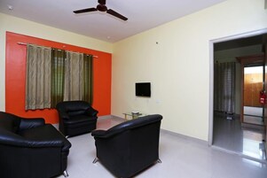 Uma sala de estar com dois sofás de couro preto, um ventilador de teto, uma TV de tela plana na parede e uma mesa de vidro.