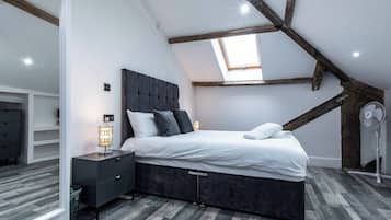 Quarto com teto inclinado, cama com cabeceira escura, mesa de cabeceira com abajur e claraboia.