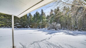 Paisagem coberta de neve com uma construção de madeira, provavelmente uma cabana ou abrigo, rodeada por densas árvores perenes.