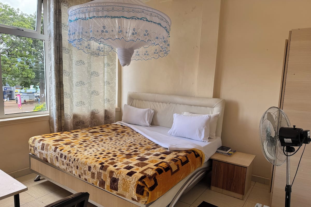 Quarto com cama, mesa de cabeceira, cadeira, ventilador e janela com cortinas.