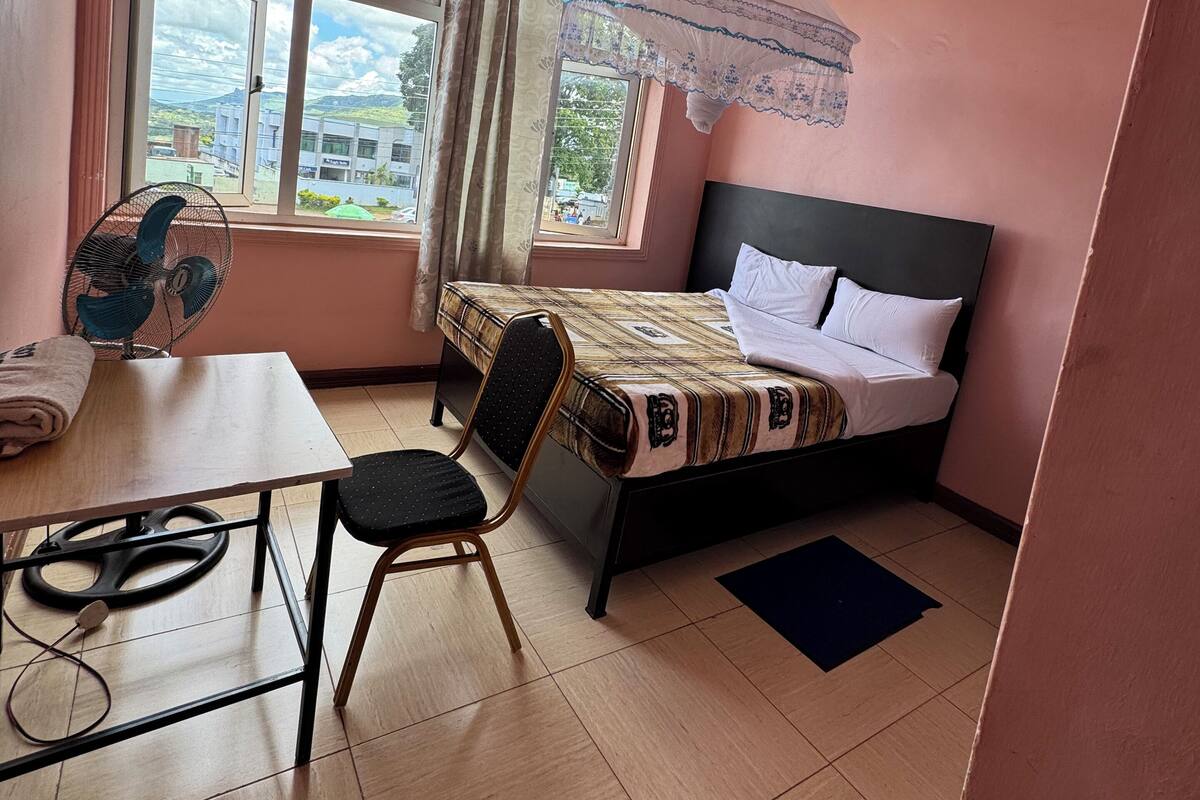 Quarto com cama, escrivaninha, cadeira, ventoinha e janela com vista para edifícios e árvores.