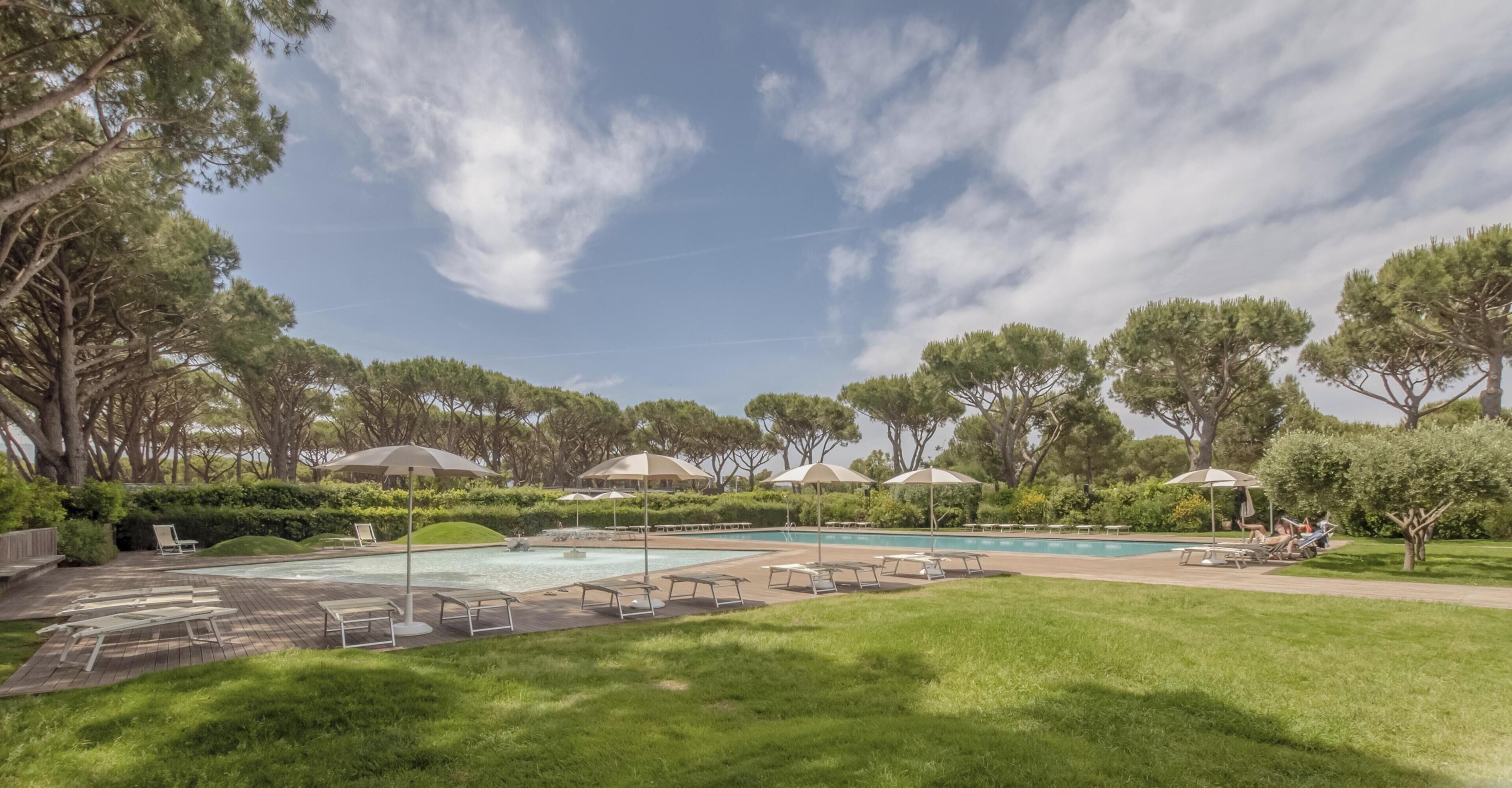 Área de piscina com espreguiçadeiras e guarda-sóis, rodeada por árvores e céu claro.