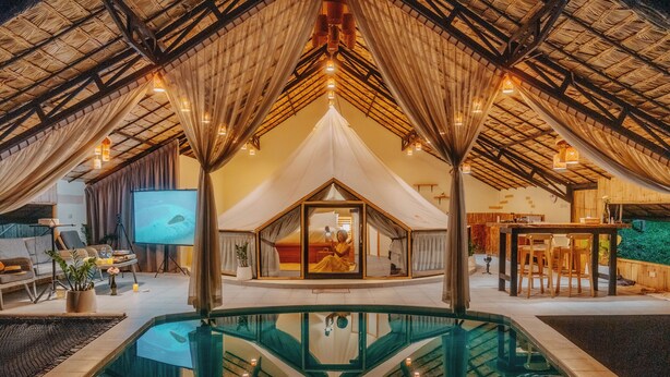 Uma área de piscina interior luxuosa, com um espaço central para sentar, uma tela grande e um ambiente acolhedor com vista para a piscina.