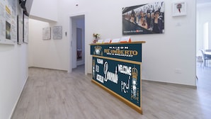 Uma receção com uma placa a indicar "MAISON du REUMBERTO BED & BREAKFAST" e uma mensagem de boas-vindas. Há quadros nas paredes e um mapa à esquerda.