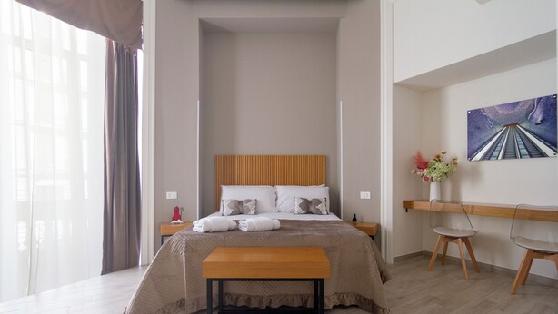 Quarto de hotel moderno com uma cama grande, cabeceira de madeira, mesa de cabeceira com luminária, um banco e uma escrivaninha com cadeiras.