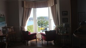 Quarto de canto com duas poltronas vermelhas, uma mesa pequena com abajur e vista para o mar através de grandes janelas.