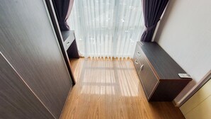 Um quarto estreito com piso de madeira, uma escrivaninha de madeira escura e uma janela com cortinas leves.