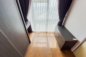 Um quarto estreito com piso de madeira, uma escrivaninha de madeira escura e uma janela com cortinas leves.