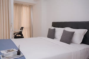 Cama bem arrumada com roupa de cama branca e cinza, cabeceira preta e mesa azul com uma revista e porta-canetas.
