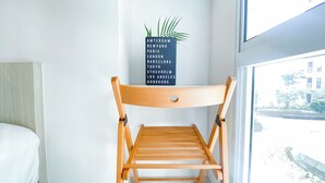 Uma cadeira de madeira num quarto claro com uma janela. Um aviso na parede lista cidades como Amsterdão, Nova Iorque e Paris.