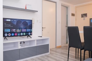 Uma sala de estar moderna com uma televisão de tela plana exibindo um programa, um móvel de entretenimento branco e uma área de jantar com cadeiras pretas.