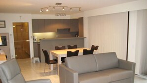 Uma sala de estar moderna com um sofá cinzento, uma mesa de jantar com cadeiras e uma área de cozinha ao fundo.