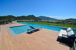 Uma área de piscina com deck de madeira, cadeiras de sol e vista para montanhas e uma cidade.