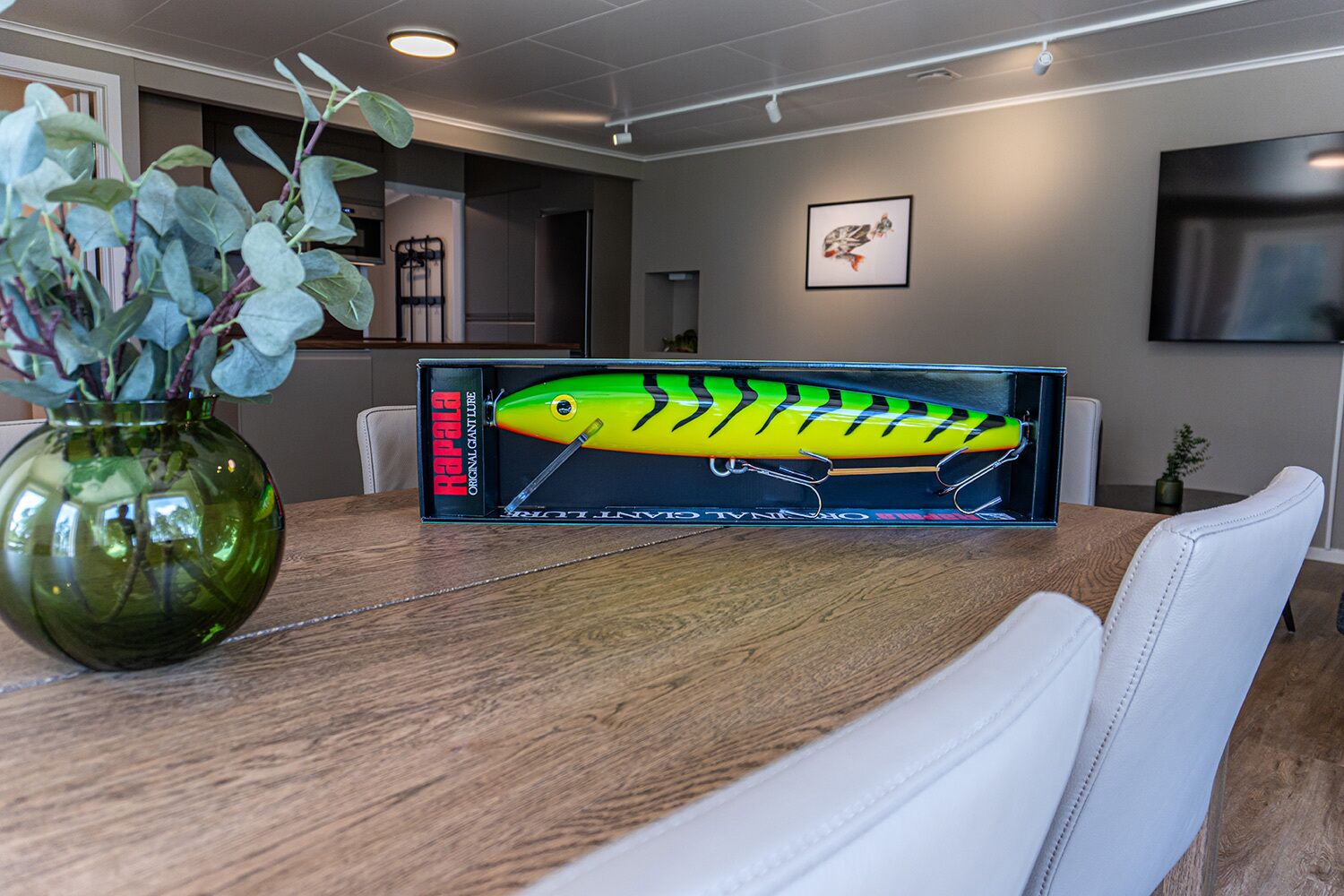 Uma mesa de jantar com um modelo de lagarto emoldurado, um vaso verde com plantas e um interior moderno com televisão e obras de arte emolduradas.