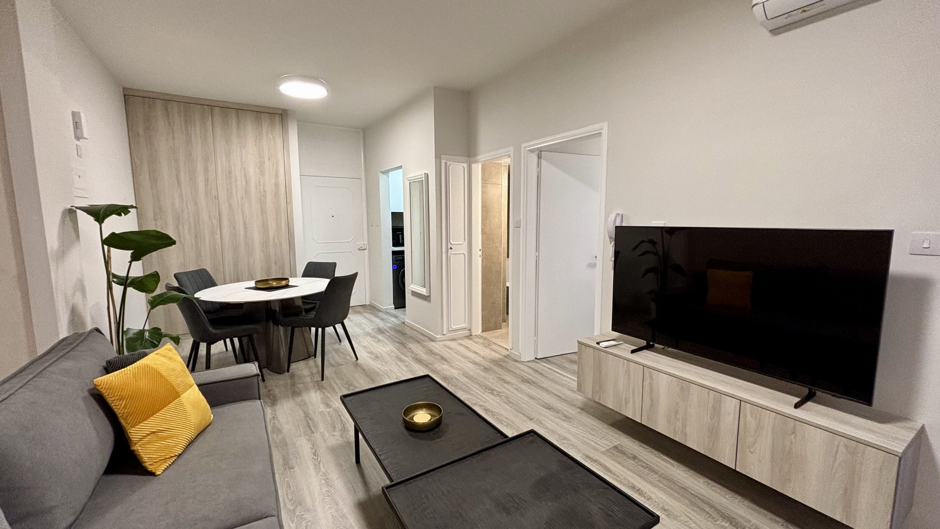 Uma sala de estar moderna com televisão de tela plana, sofá cinza e um espaço de jantar com mesa redonda e cadeiras.
