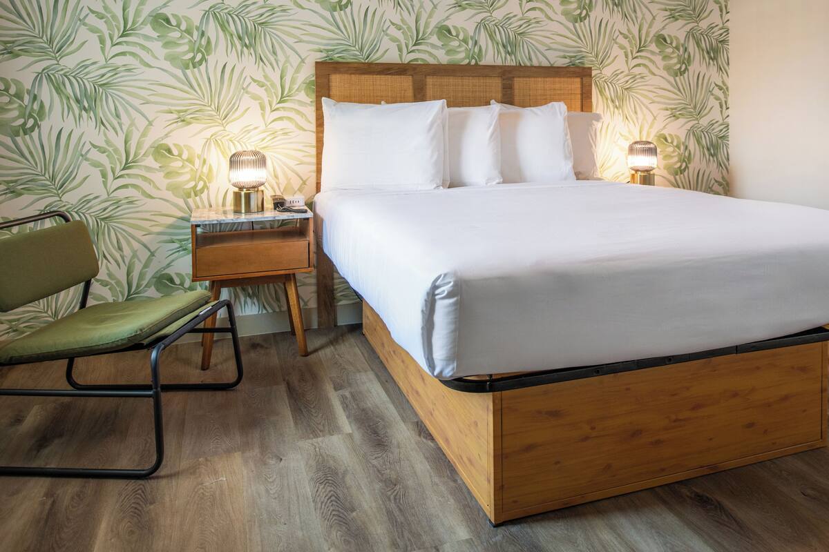 Um quarto bem arrumado com uma cama de madeira, uma cadeira verde, uma mesa de cabeceira com abajur e uma parede com padrão de folhas.