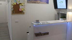 Balcão de recepção com placa de "Welcome", um mural de cortiça com anotações, uma fotografia emoldurada de paisagem e uma tela de TV exibindo uma mensagem de boas-vindas.
