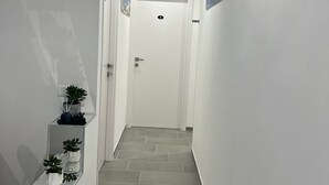 Um corredor com piso ladrilhado, paredes brancas e quadros emoldurados. Há uma porta com o número 8 e um pequeno armário branco com plantas à esquerda.