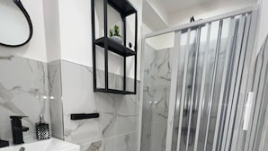 Casa de banho moderna com lavatório branco, estantes pretas, cabine de duche em vidro e paredes em mármore.