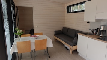 Espaço compacto com mesa de jantar, cadeiras, sofá, pequena cozinha com pia e cafeteira, e janela com vista para árvores.