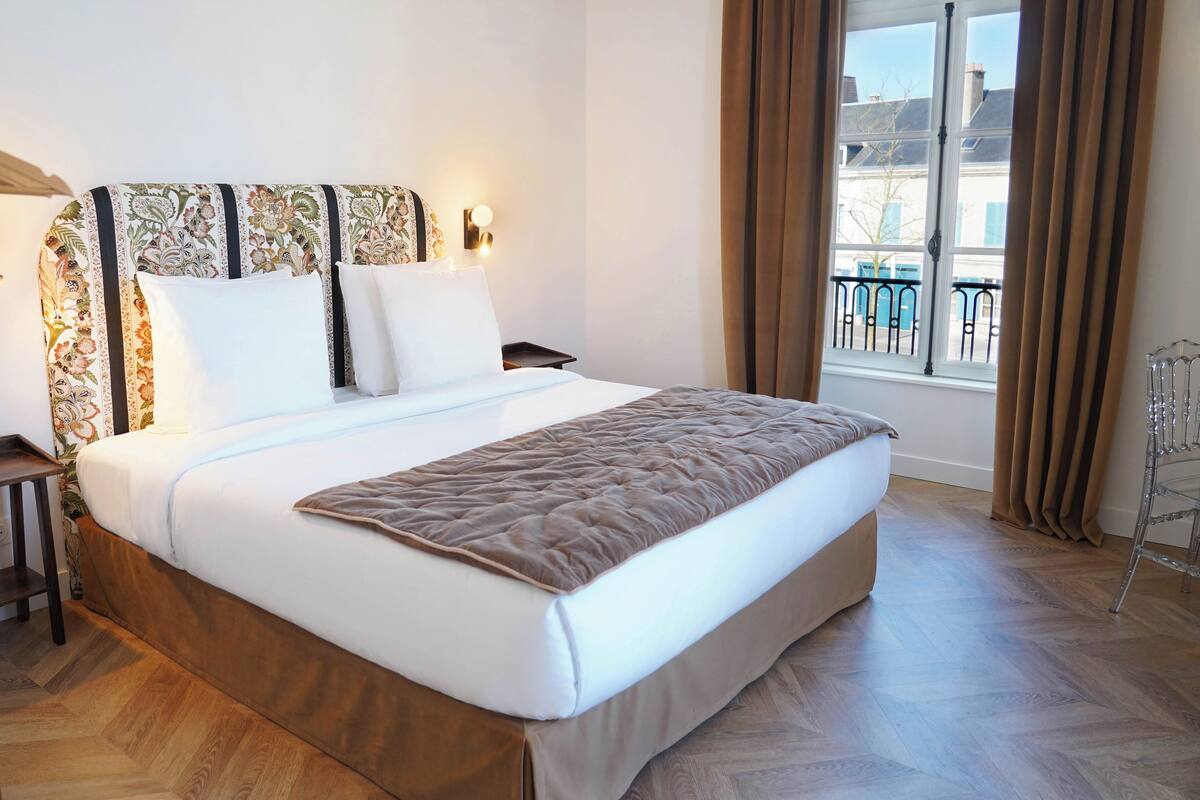 Cama bem arrumada com cabeceira estampada, mesa de cabeceira com abajur e vista para a varanda através de uma janela.