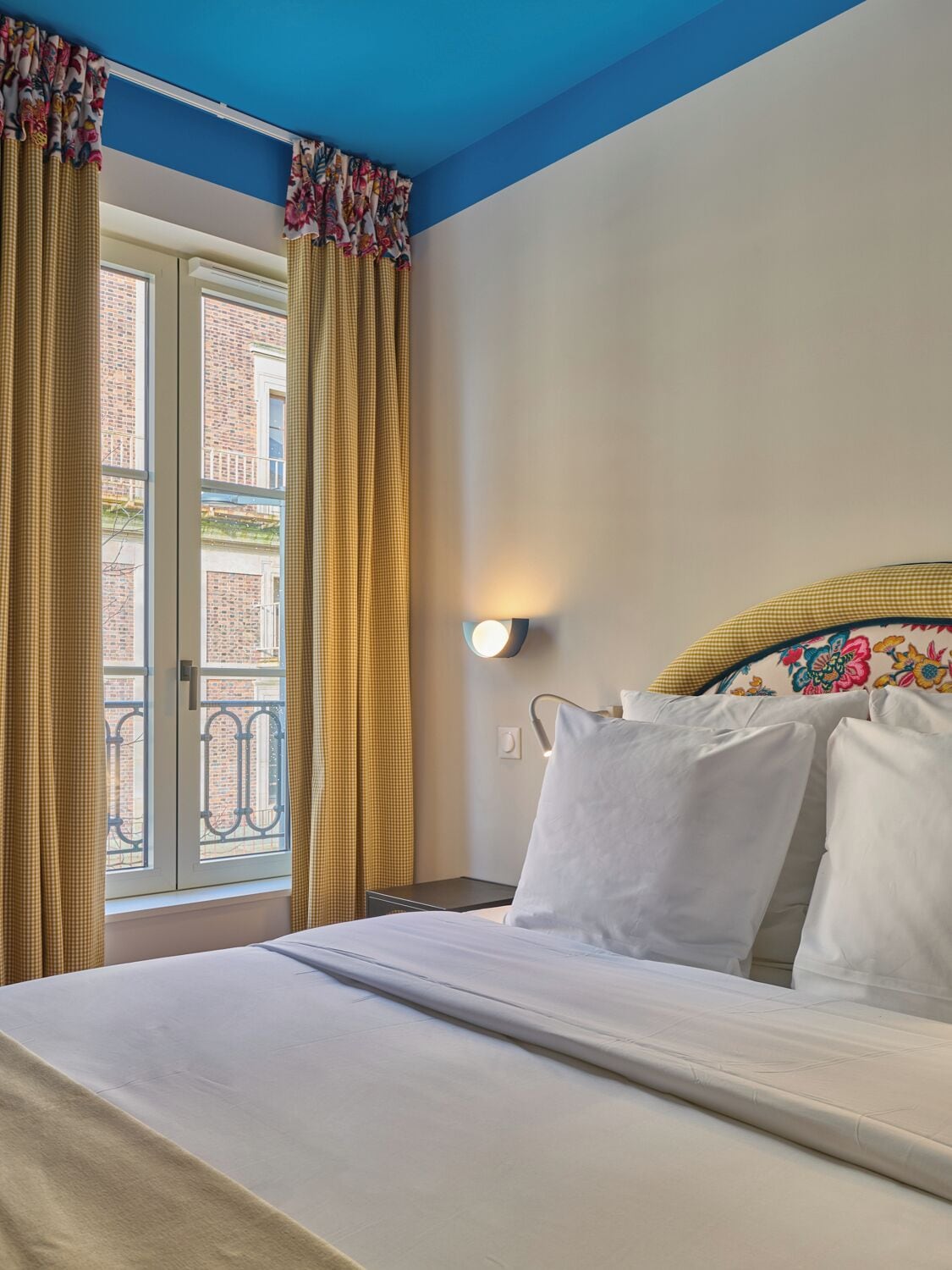 Quarto de hotel com cama, cortinas, luminária fixada na parede e vista para um edifício de tijolos pela janela.