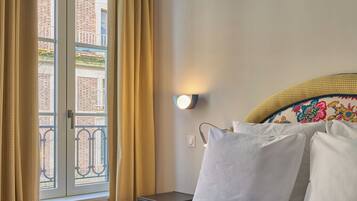 Quarto de hotel com cama, cortinas, luminária fixada na parede e vista para um edifício de tijolos pela janela.