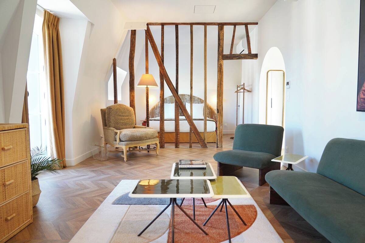 Sala moderna com sofá, poltrona e mesa de centro. Uma cama com cabeceira de madeira pode ser vista ao fundo.
