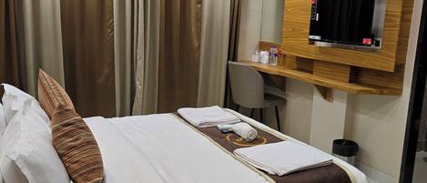 Cama bem arrumada com roupa de cama branca e castanha, cabeceira de madeira e televisão montada na parede acima.