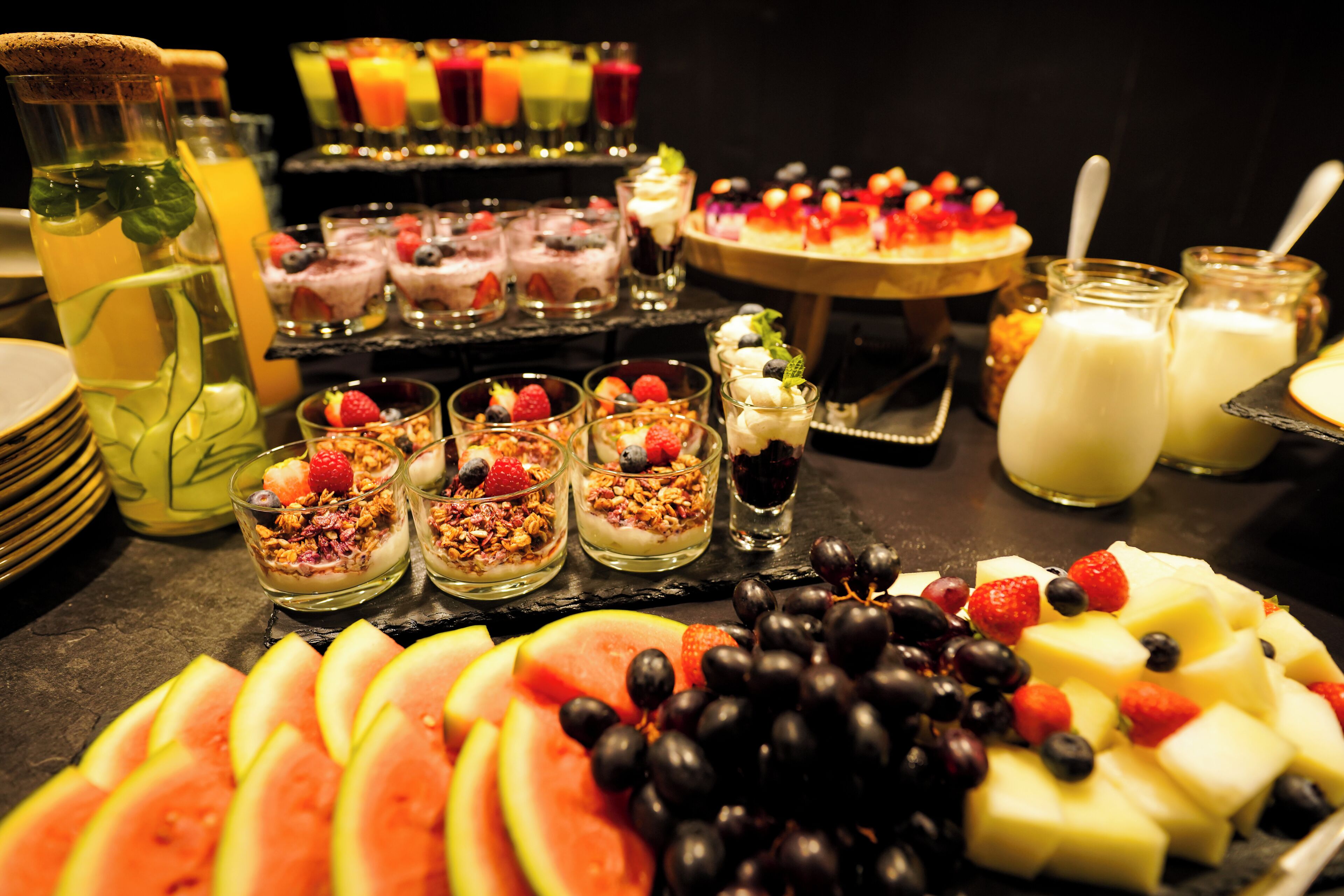 Diversos alimentos e bebidas sobre uma mesa, incluindo frutas, potes de iogurte e uma seleção de sucos.