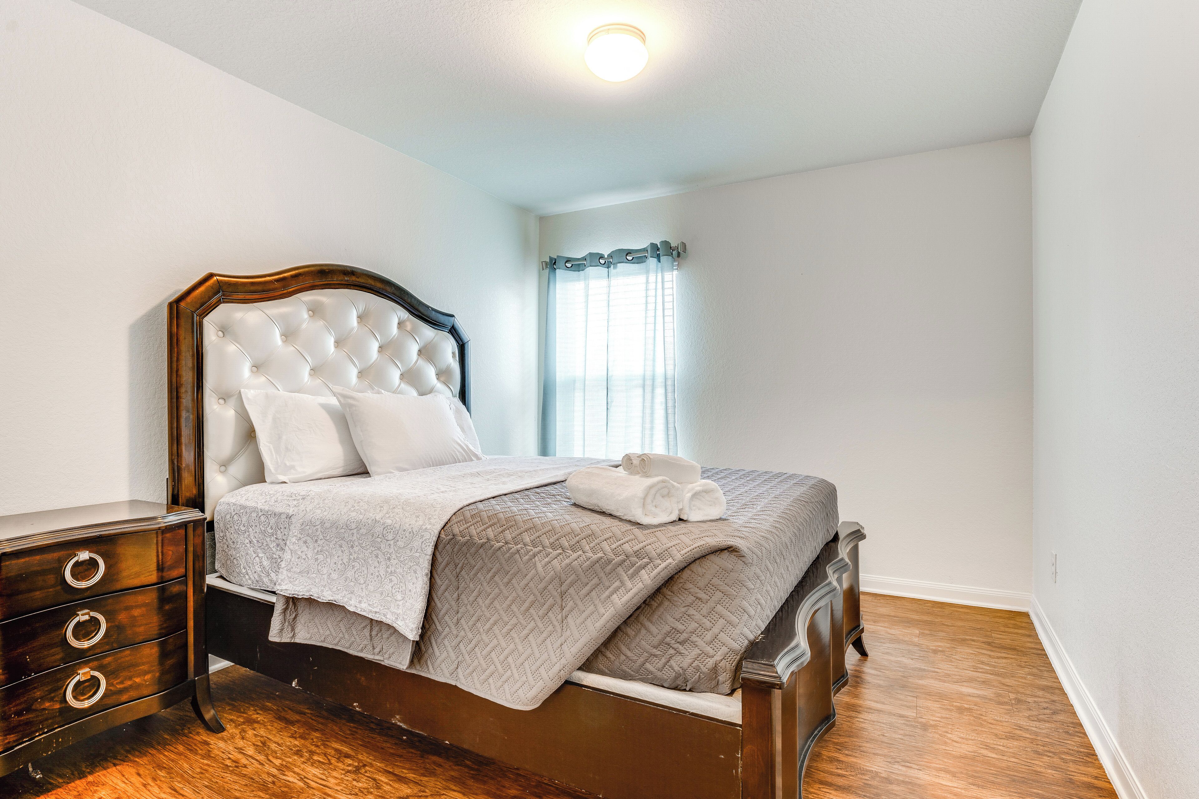 Uma cama bem arrumada com cabeceira estofada, uma mesinha de cabeceira de madeira com gavetas e uma janela com cortinas.