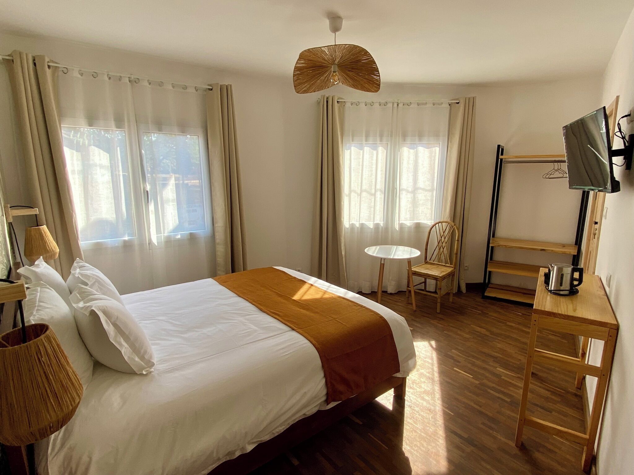 Quarto de hotel com cama, uma mesinha, uma cadeira, televisão e janela com cortinas.