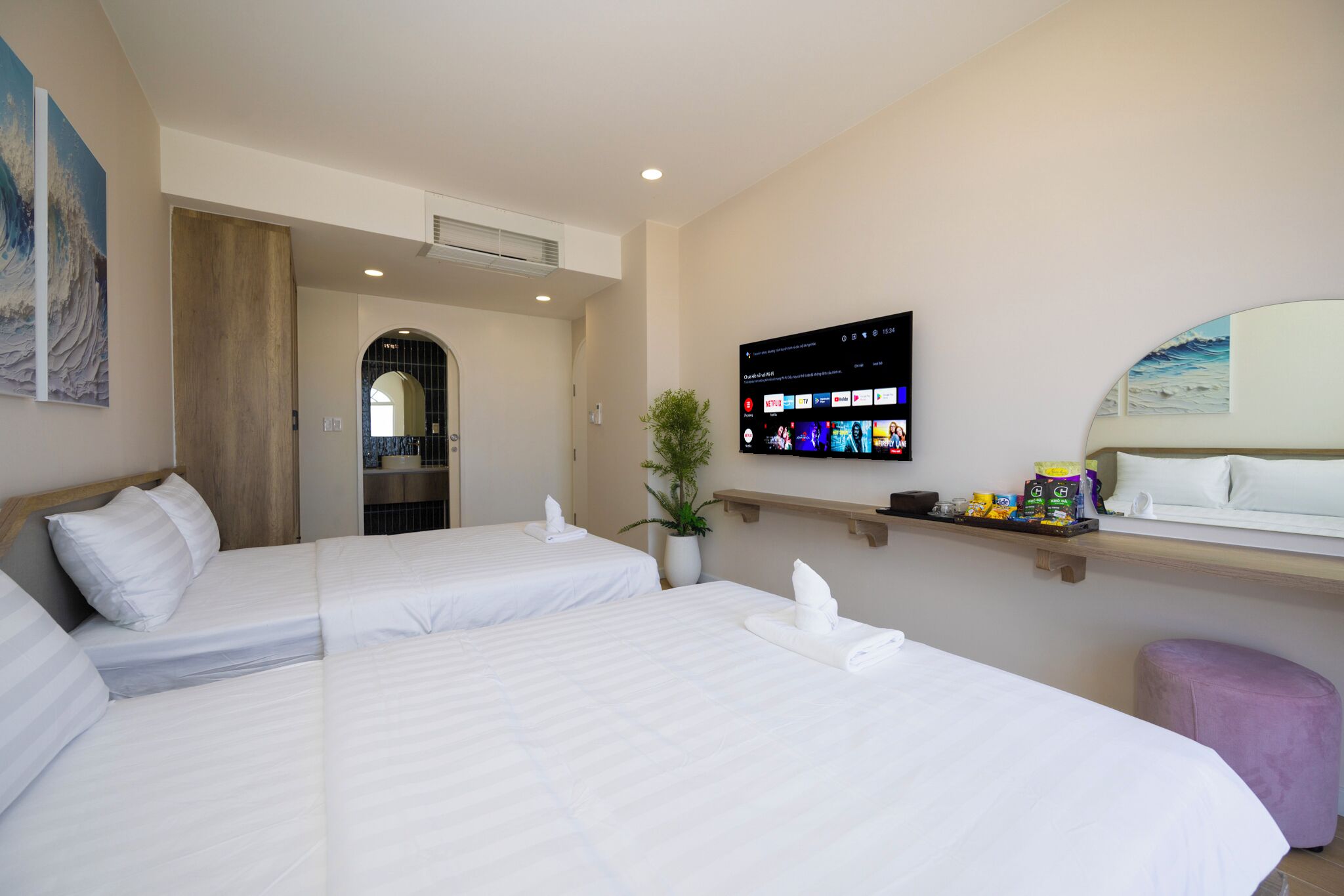 Quarto de hotel com duas camas, uma televisão fixada na parede, uma pequena prateleira com lanches e uma planta em vaso.