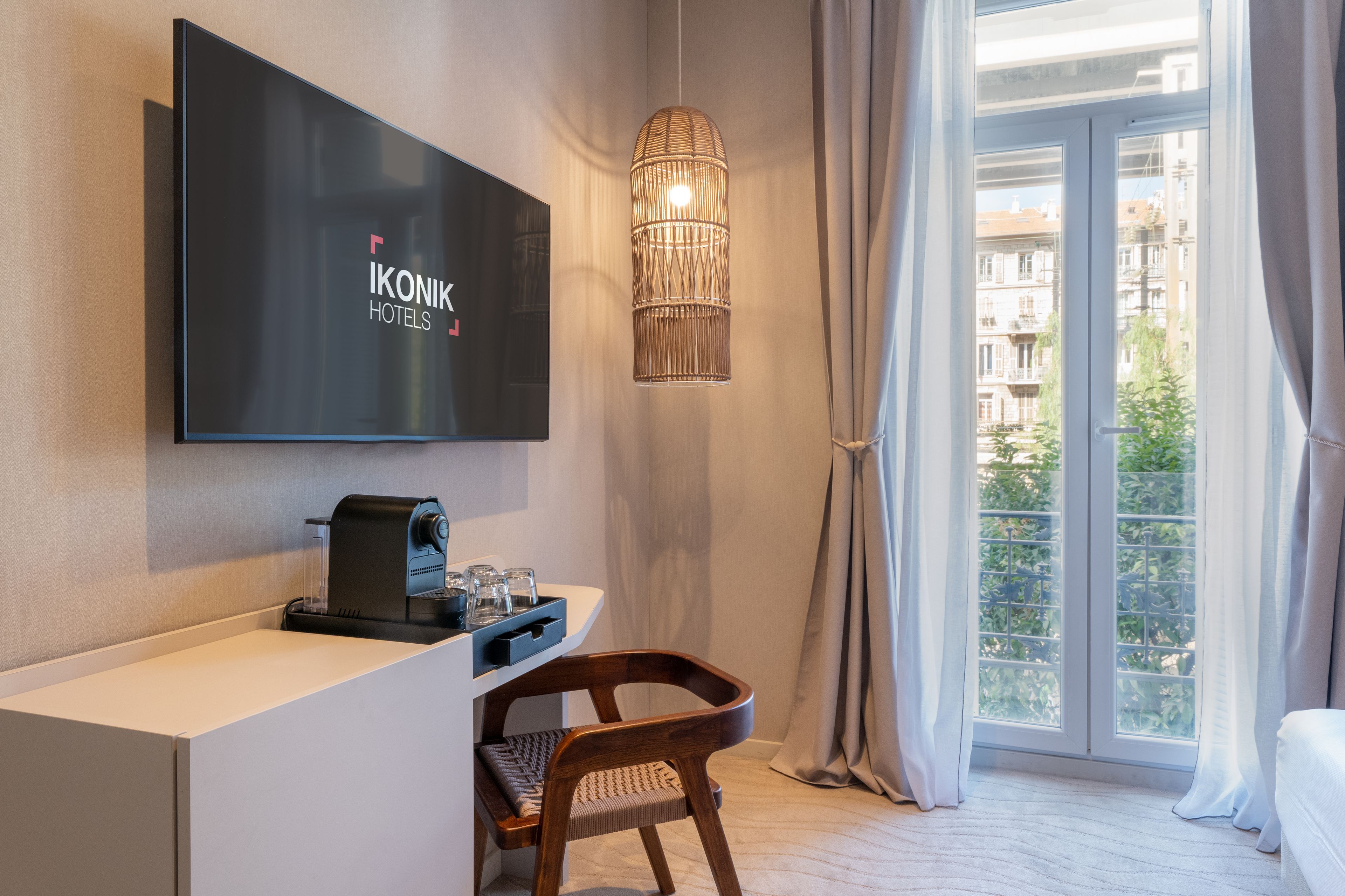 Quarto de hotel com televisão de tela plana na parede, máquina de café no balcão, luminária pendente de vime, cadeira de madeira e uma grande janela com cortinas.
