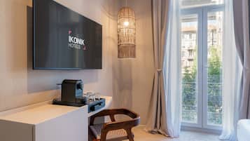 Quarto de hotel com televisão de tela plana na parede, máquina de café no balcão, luminária pendente de vime, cadeira de madeira e uma grande janela com cortinas.