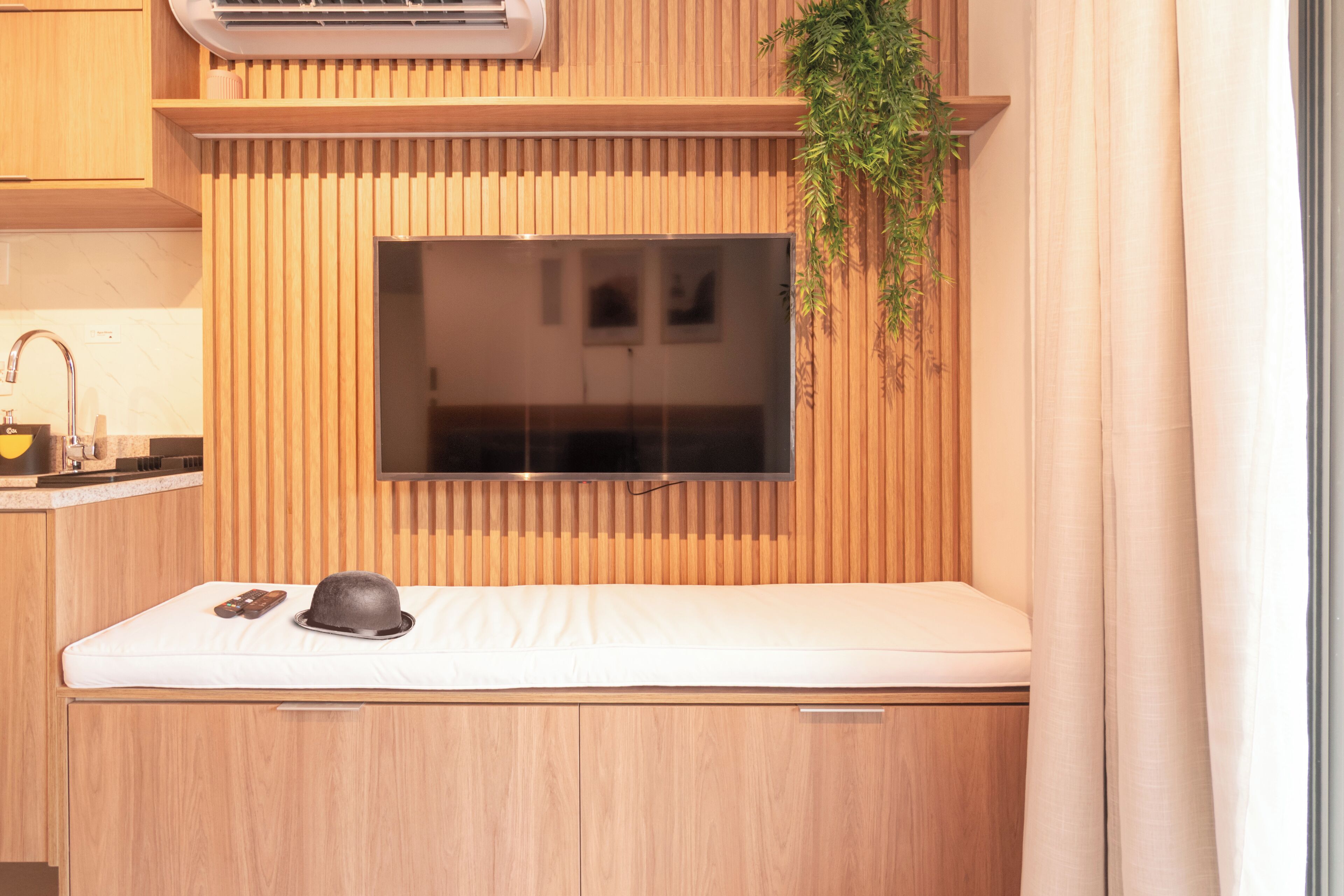 Quarto compacto com televisão integrada, um pequeno espaço para sentar e janela com persianas.