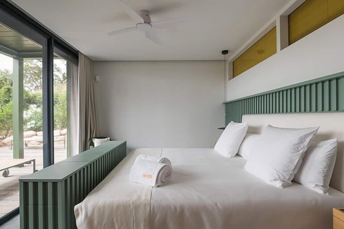 Quarto de hotel moderno com uma cama grande, um ventilador de teto e vista para o exterior através de uma porta de correr de vidro.