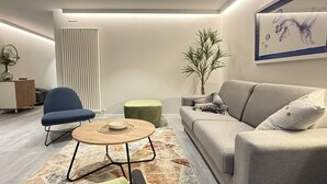 Uma sala de estar moderna com um sofá cinzento, uma poltrona azul, uma mesa de centro redonda e um banquinho verde.