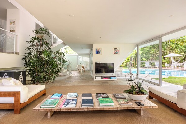 Uma sala de estar moderna com uma televisão grande, uma mesa de centro com livros, um sofá e vista para uma piscina.