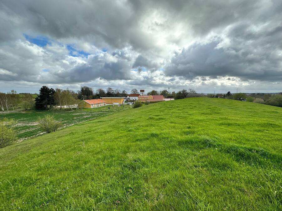 Paisagem rural com um amplo campo verde, um edifício com telhado vermelho e céu nublado.