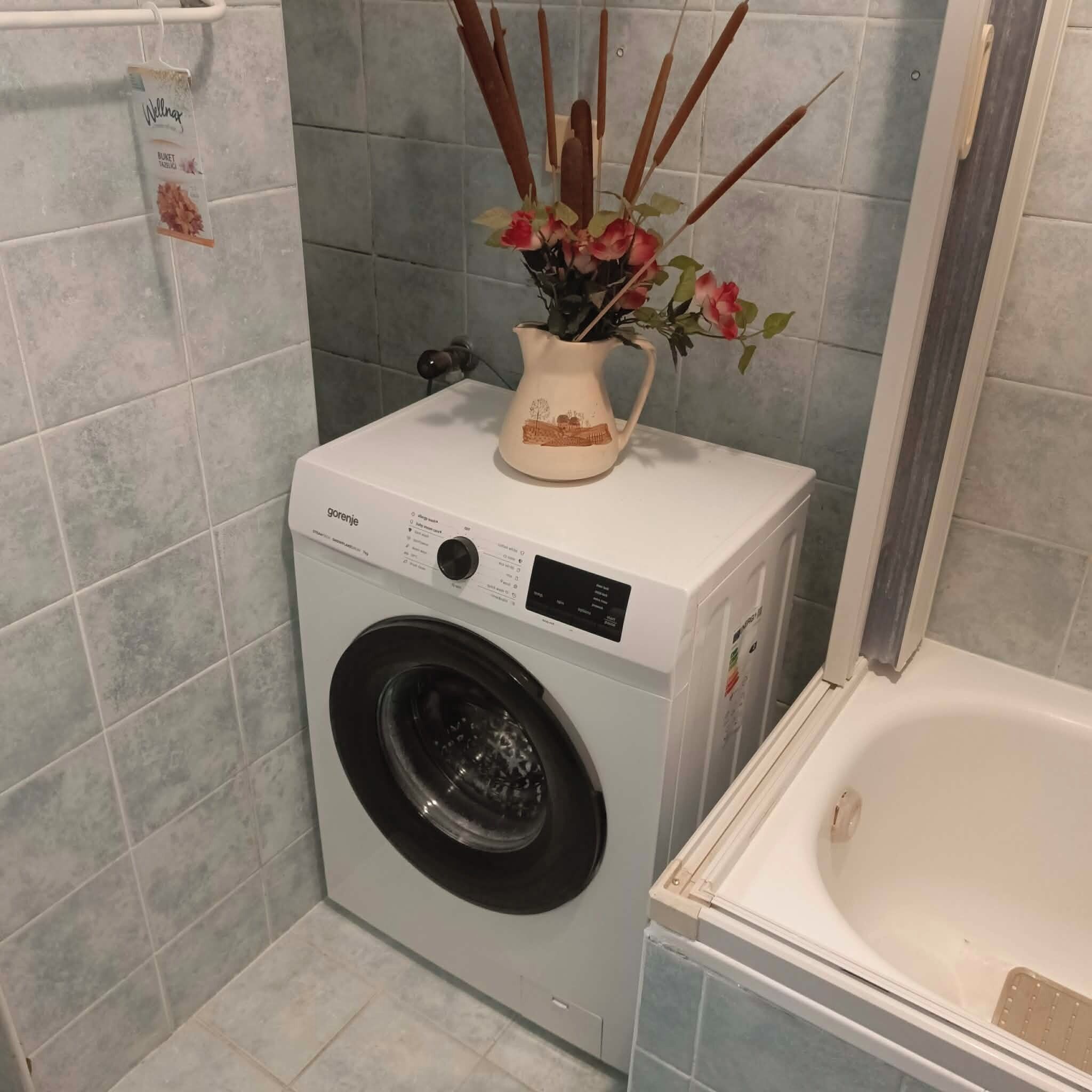 Máquina de lavar compacta com painel de controle, parede revestida de azulejos brancos e um pequeno vaso com flores em cima.
