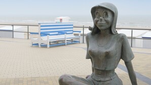 Estátua de mulher com chapéu e vestido, um banco listrado em azul e branco, e uma praia ao fundo.