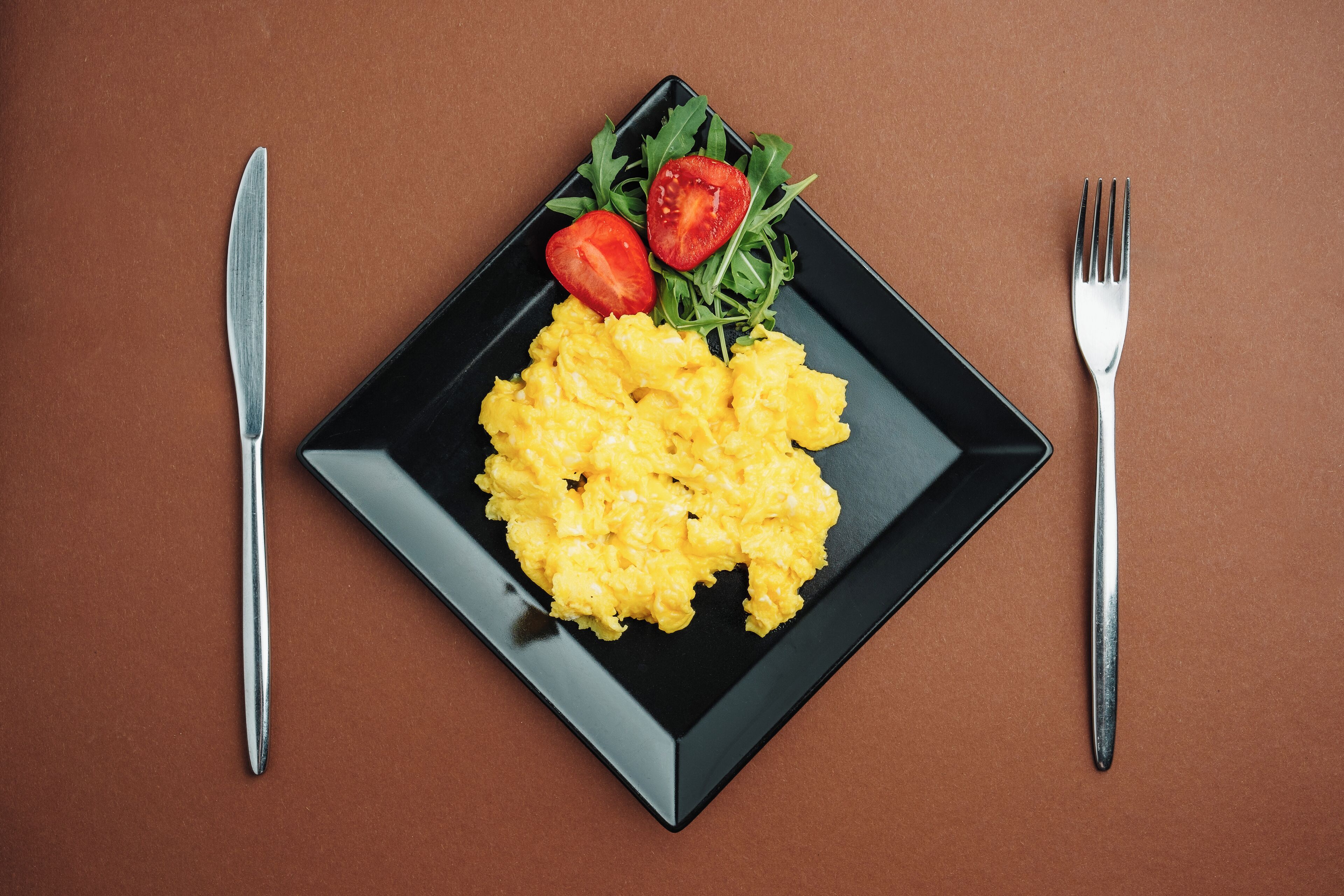 Um prato preto com ovos mexidos, duas fatias de tomate e uma salada verde sobre uma mesa marrom.