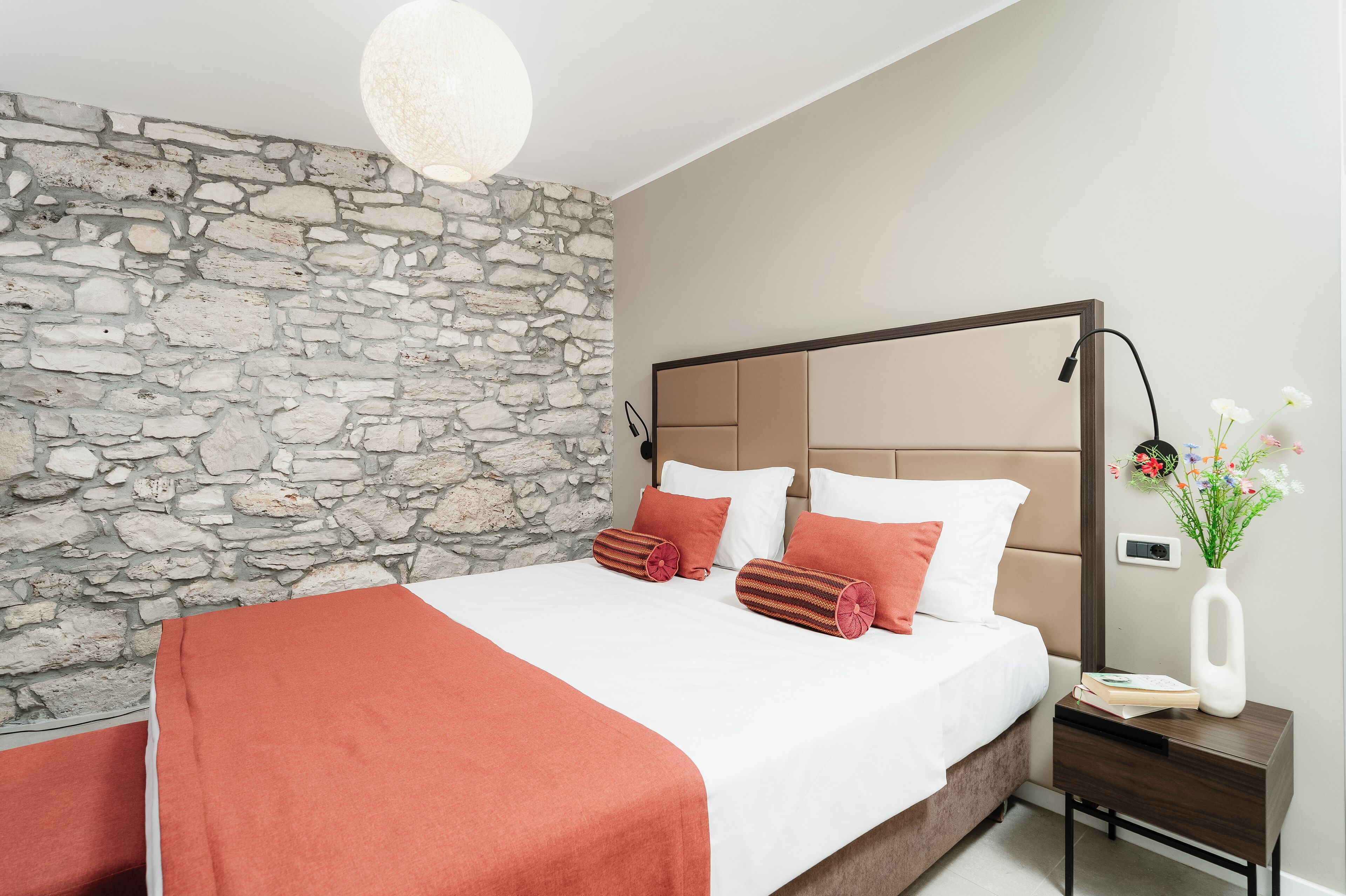 Quarto com parede de pedra, cama com cabeceira de madeira, mesa de cabeceira, luminária e um vaso com flores.