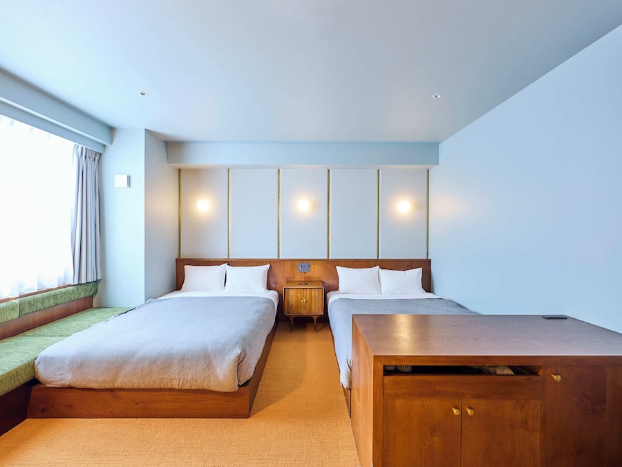 Quarto de hotel com duas camas, uma mesa de cabeceira de madeira e uma escrivaninha.