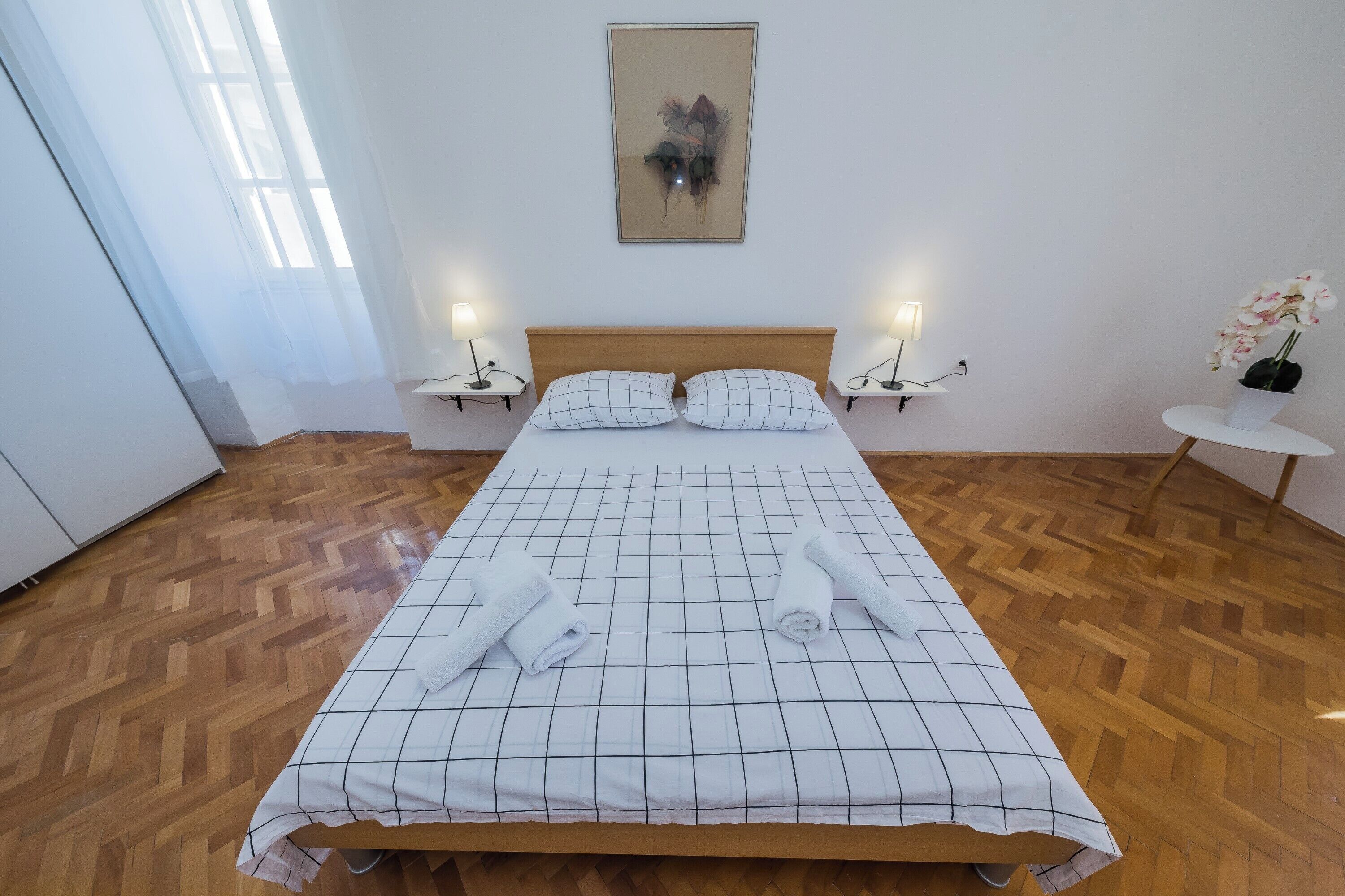 Cama bem arrumada com colcha xadrez e dois rolos de toalha, ladeada por mesas de cabeceira com abajures, em quarto com piso de madeira e uma janela grande com cortinas leves.