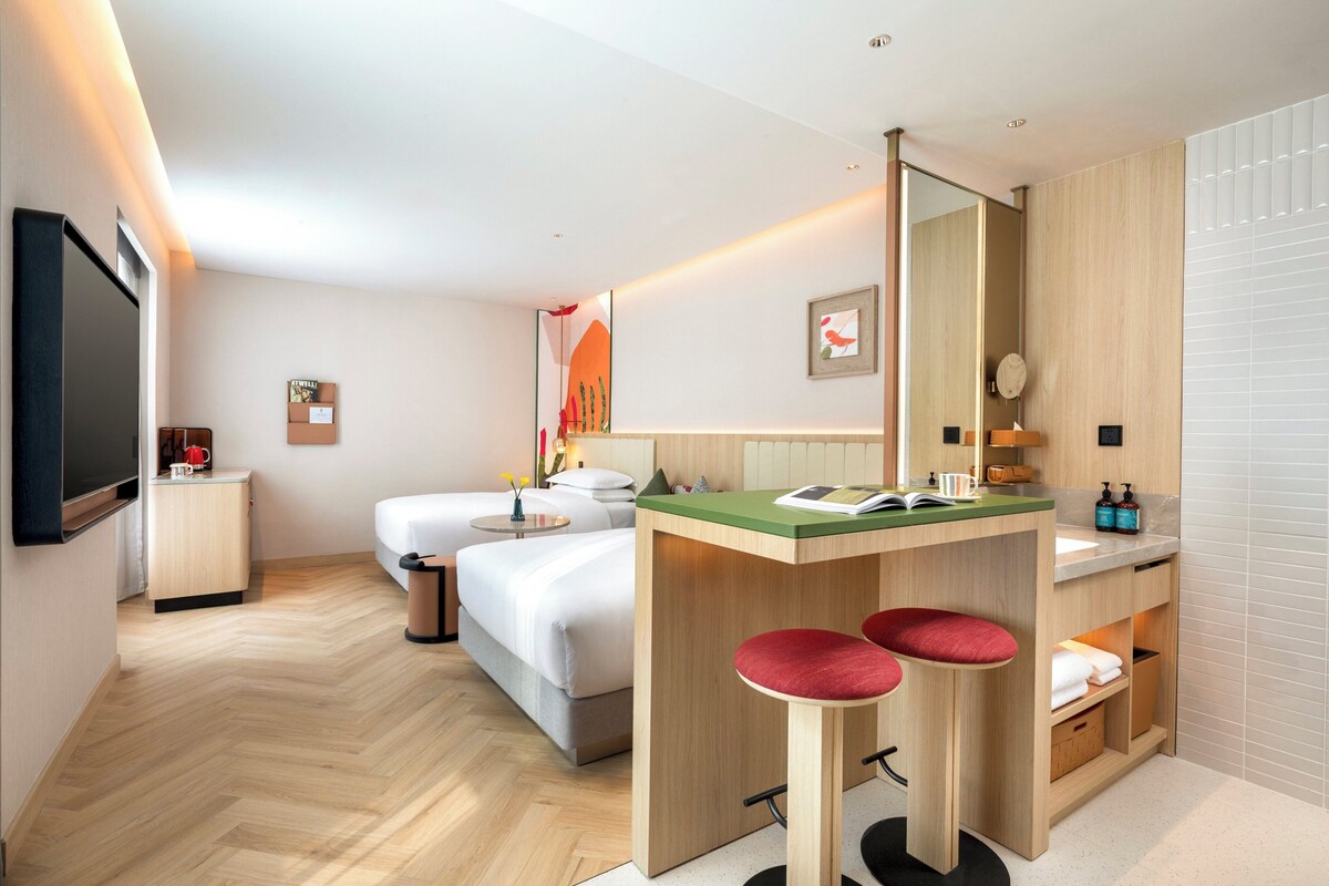 Quarto de hotel moderno com uma cama, uma escrivaninha com dois banquetes vermelhos, um espelho, uma TV fixada na parede e uma pequena mesa de cabeceira com uma luminária.