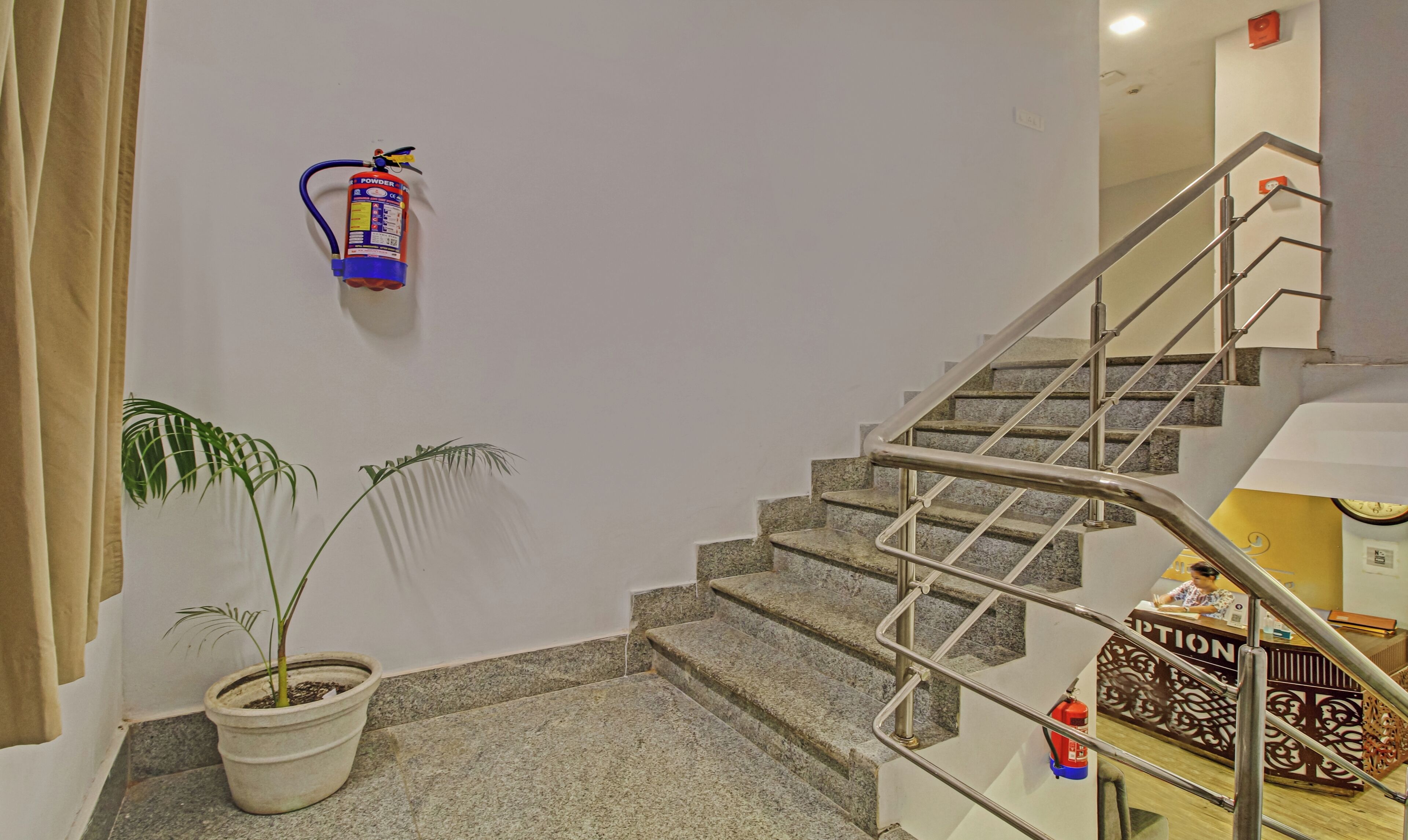 Uma escada com guarda-corpos de metal, um extintor fixado na parede e um vaso de planta no patamar.